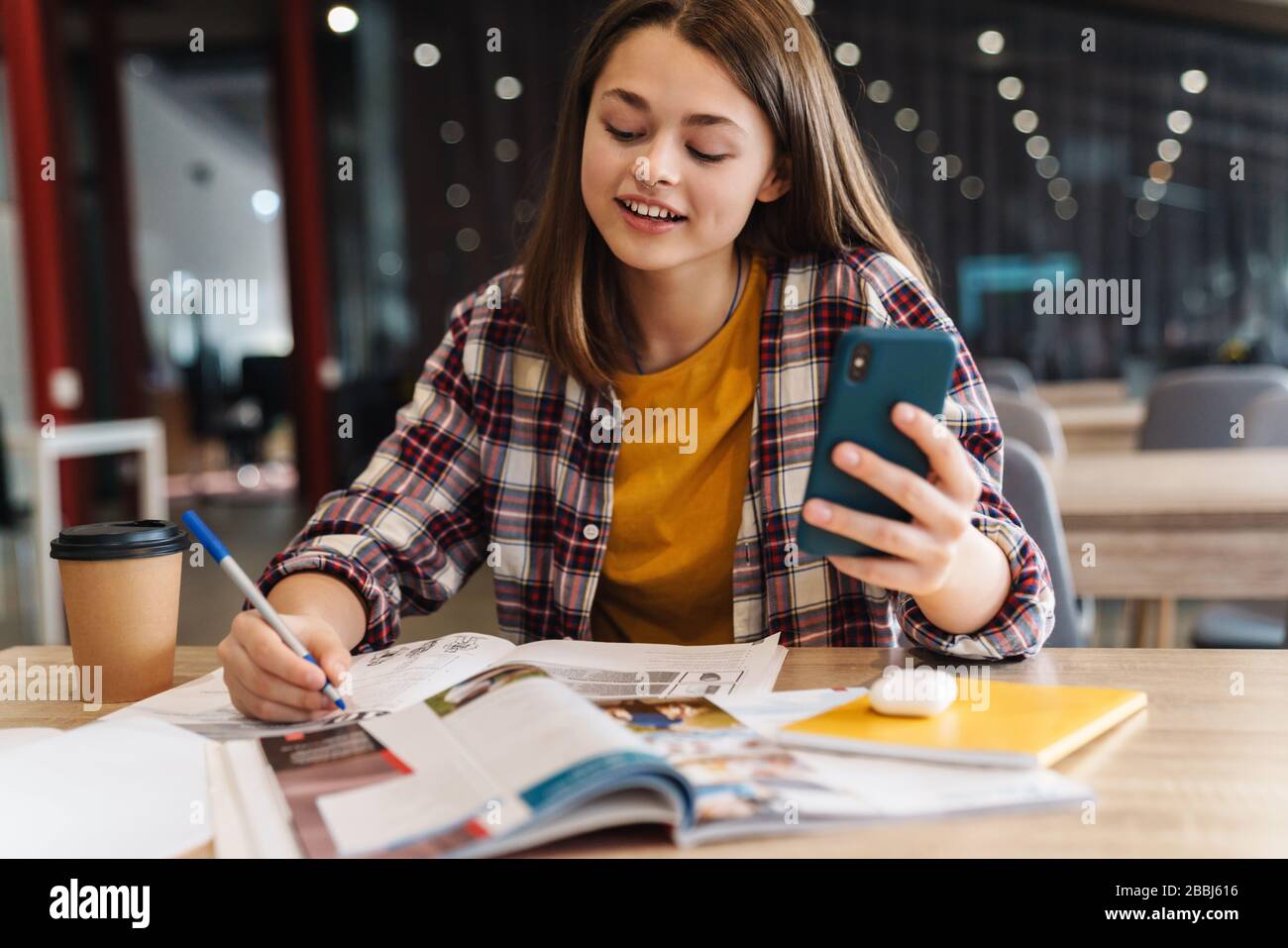 Image of joyful beautiful girl using cellphone while doing homework ...