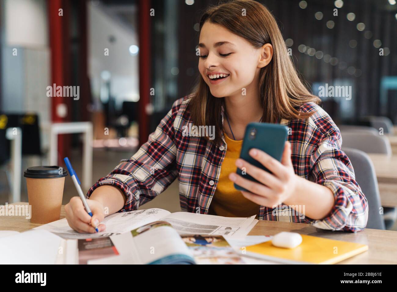 Image of joyful beautiful girl using cellphone while doing homework ...