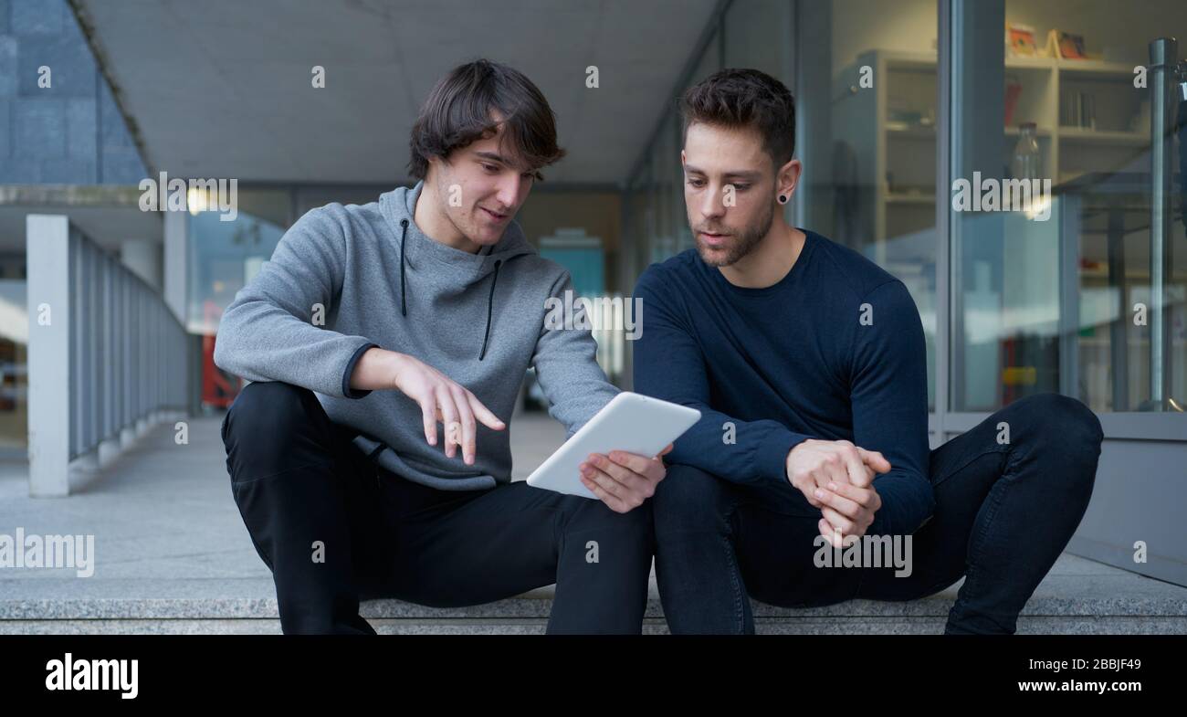 Front view of two young men talking sitting on a city staircase and lo ...