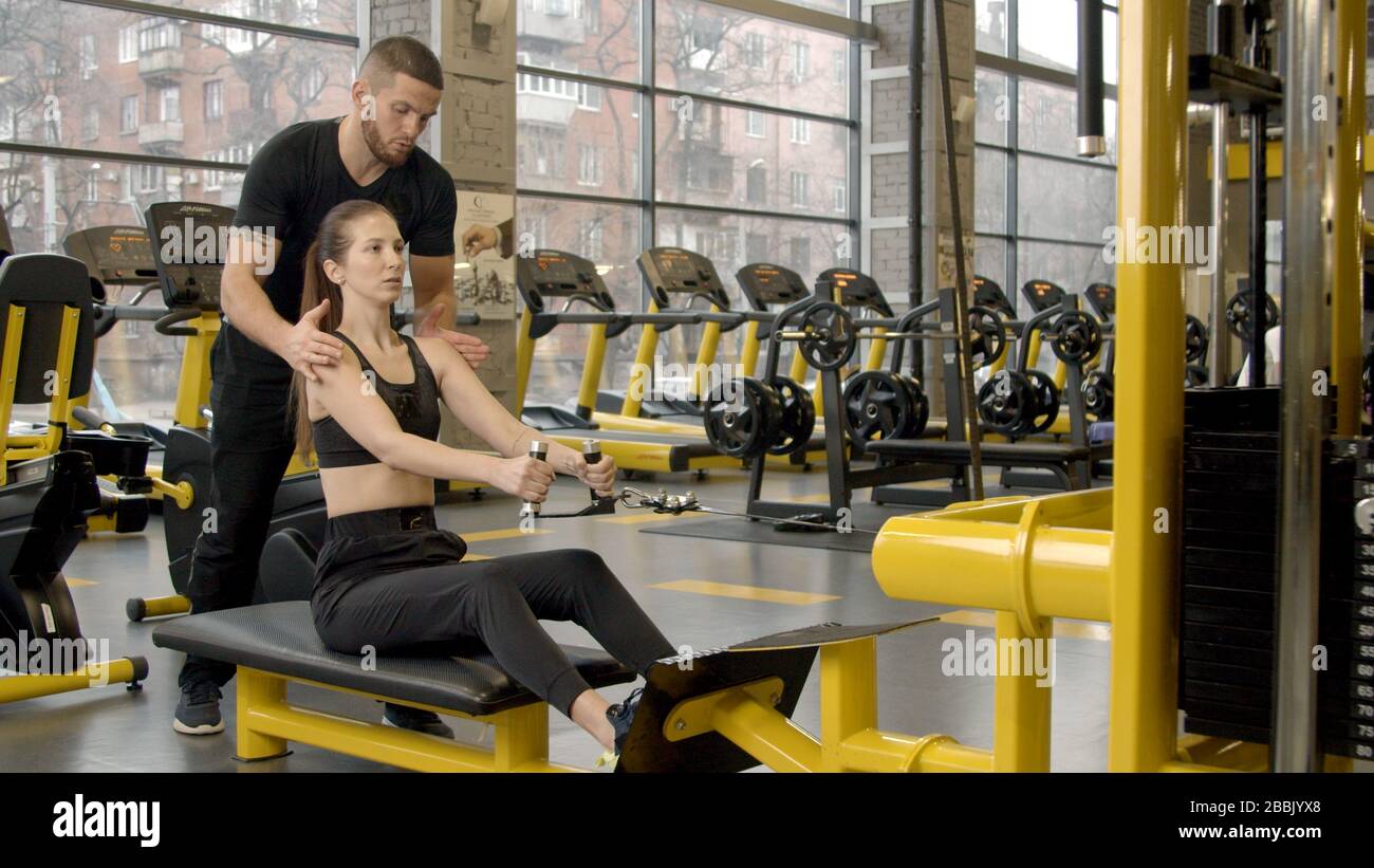 Young woman with instructor working out in gym by using rowing machine ...