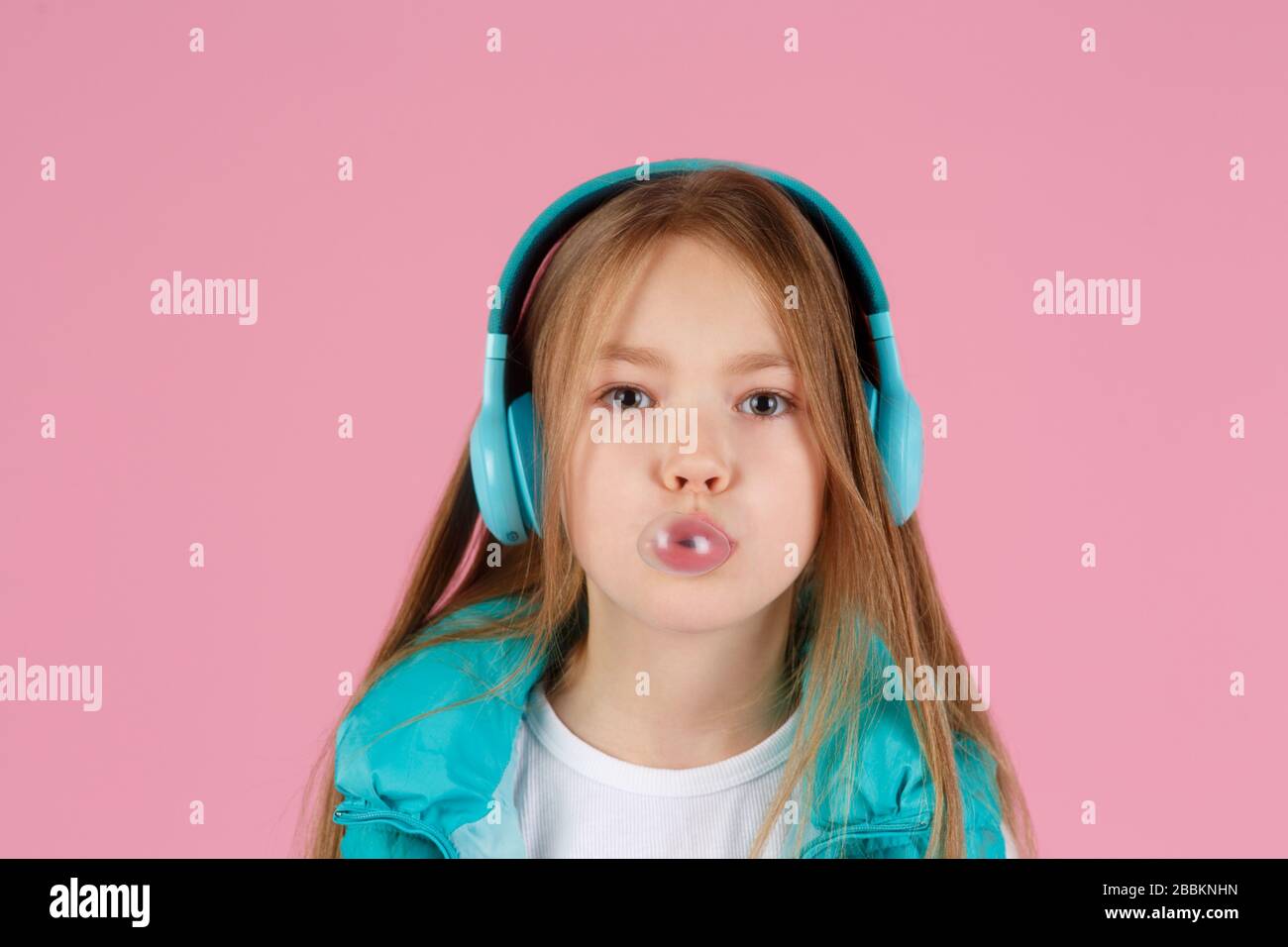A little girl with headphones explodes pink chewing gum on a pink ...