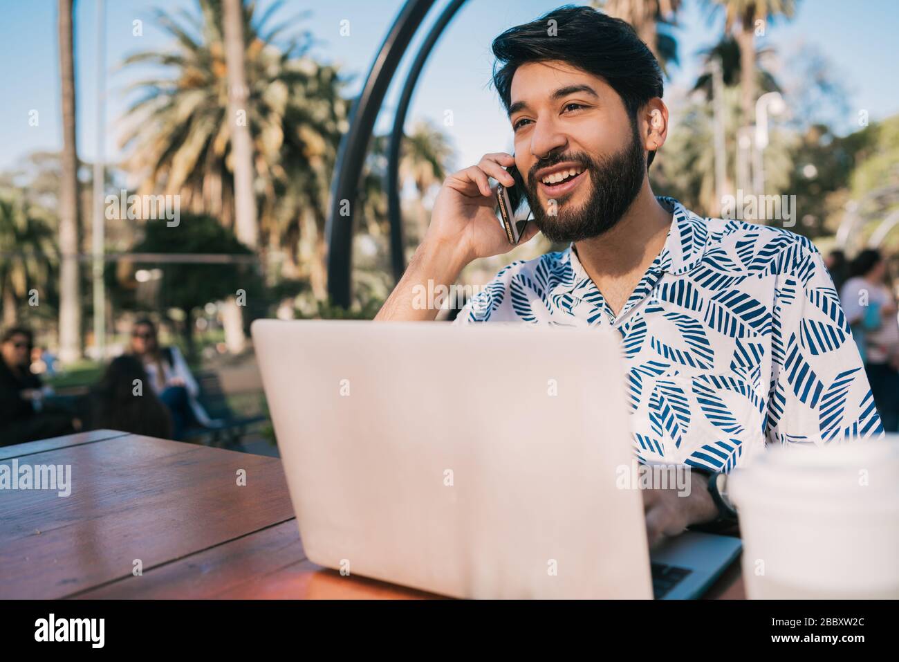 Portrait of young man using his laptop and talking on the phone while ...