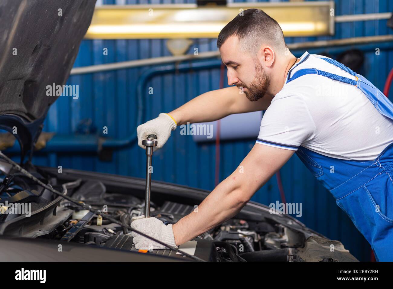Handsome young male auto mechanic in special uniform clothes tightens ...