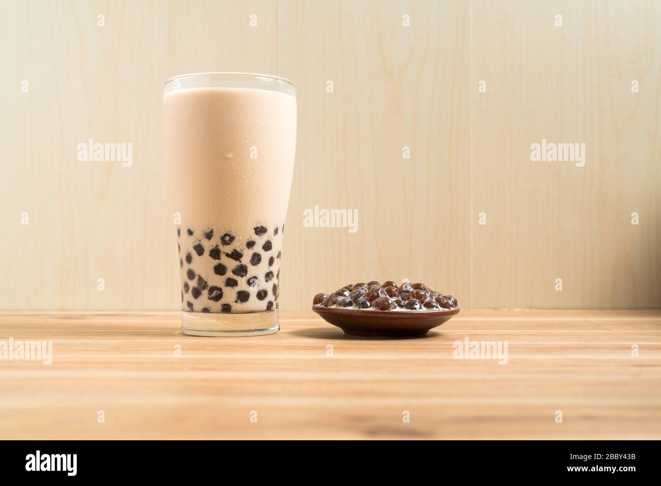 A glass cup of pearl milk tea (also called bubble tea) and a plate of tapioca ball on wooden ...