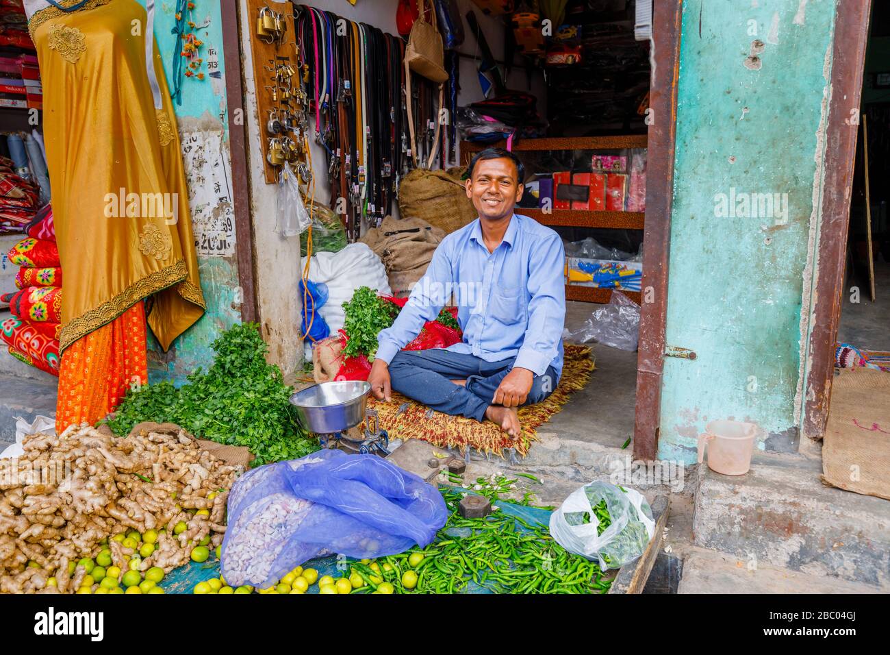 Shopkeeper sitting in the doorway of a small shop selling vegetables ...