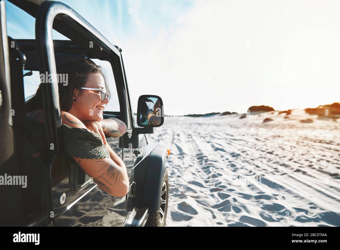 Smiling young woman leaning out jeep window, enjoying beach road trip ...