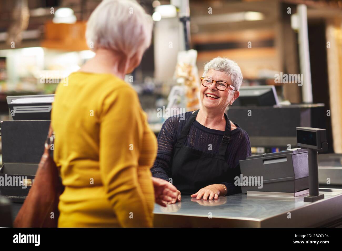 Happy senior female cashier greeting customer at supermarket checkout ...