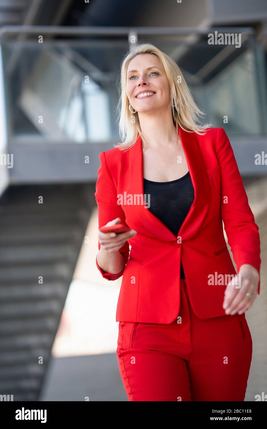 Blond smiling businesswoman wearing red suit and holding smartphone ...