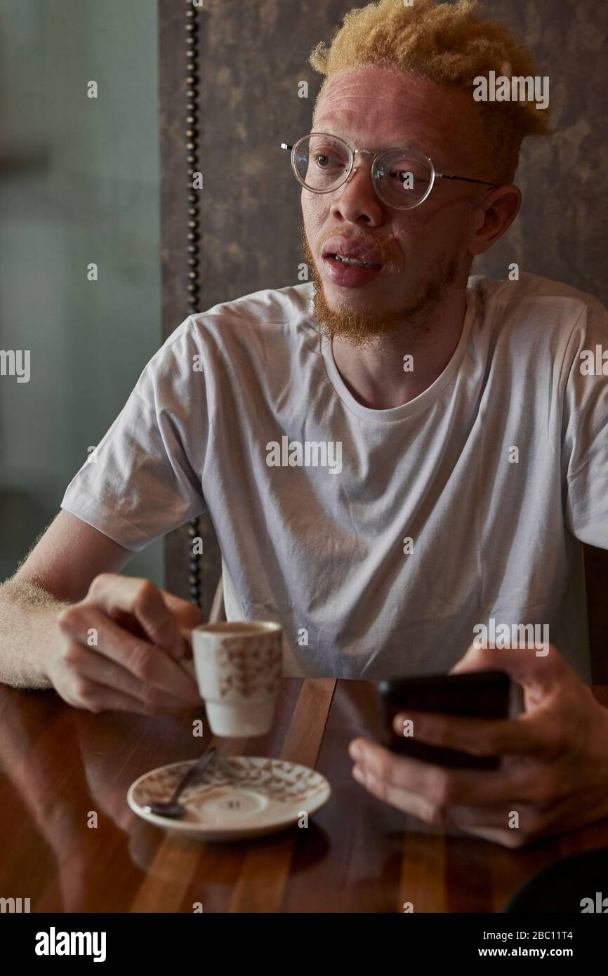 Albino man with round glasses having a coffee and checking his phone in ...