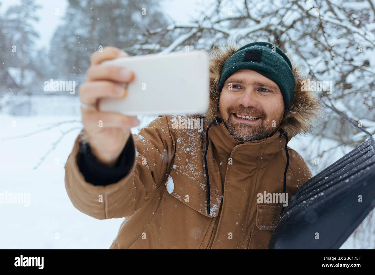 Portrait of smiling man with snow shovel taking selfie with cell phone ...