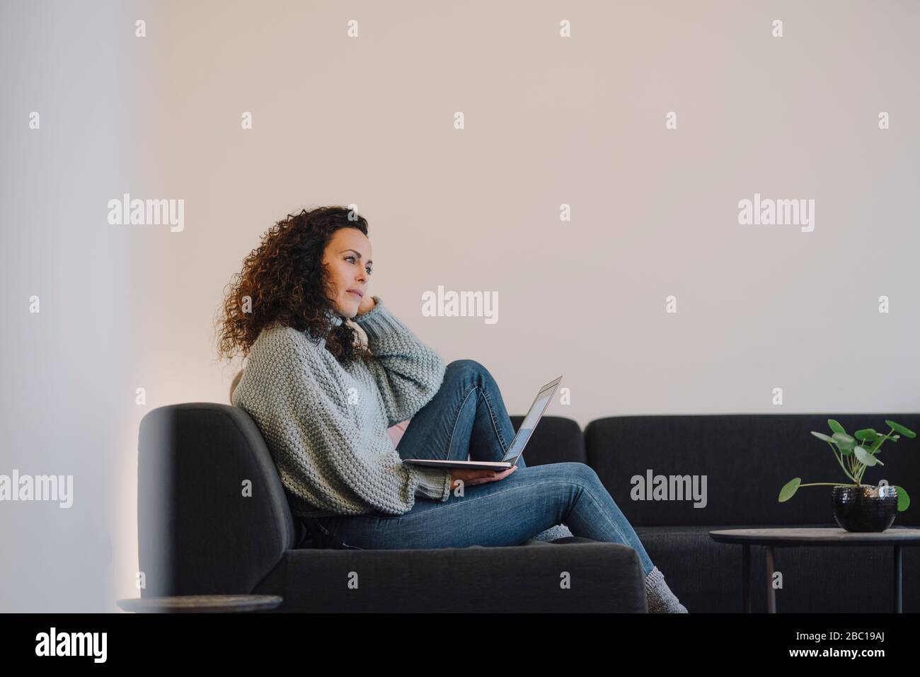 Woman sitting on couch, using laptop, thinking Stock Photo - Alamy