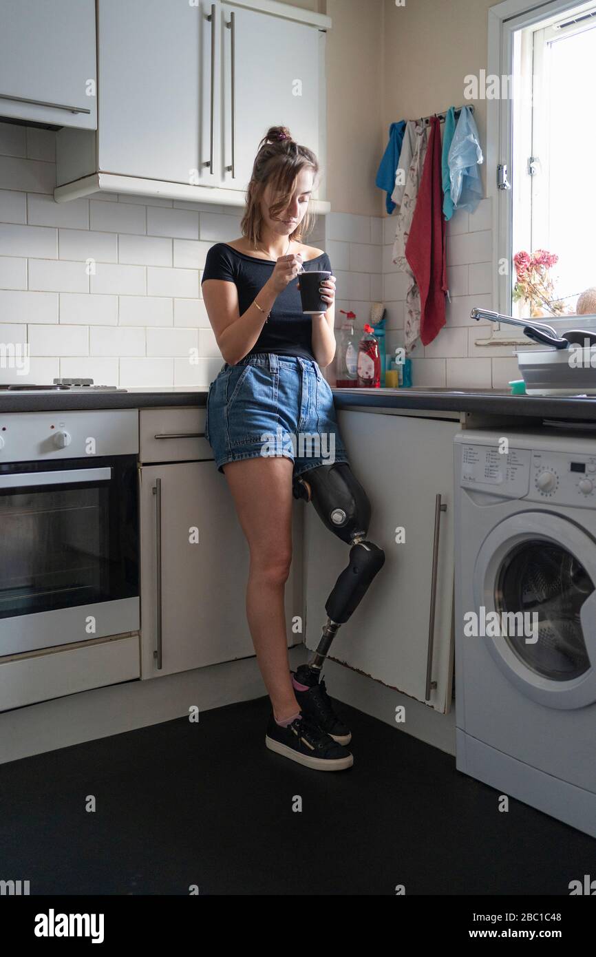 Young woman with leg prosthesis drinking coffee in kitchen at home ...