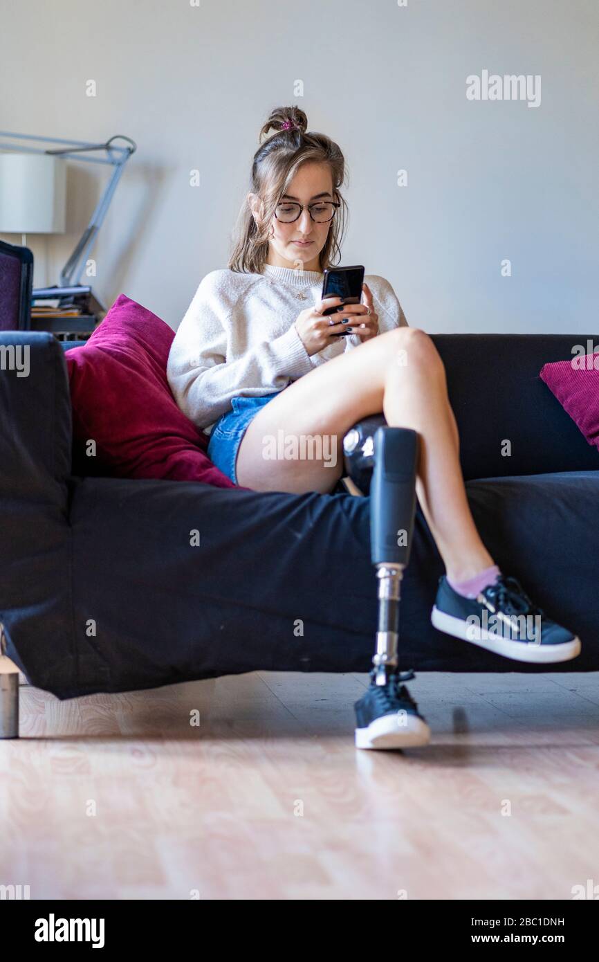 Young woman with leg prosthesis sitting on couch at home using ...