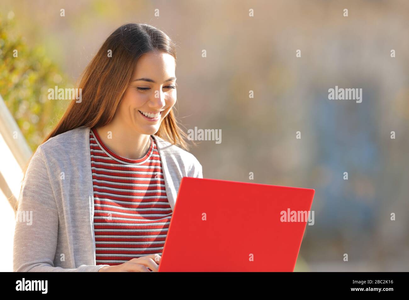 Happy girl using a red laptop sitting outdoors in a university campus ...