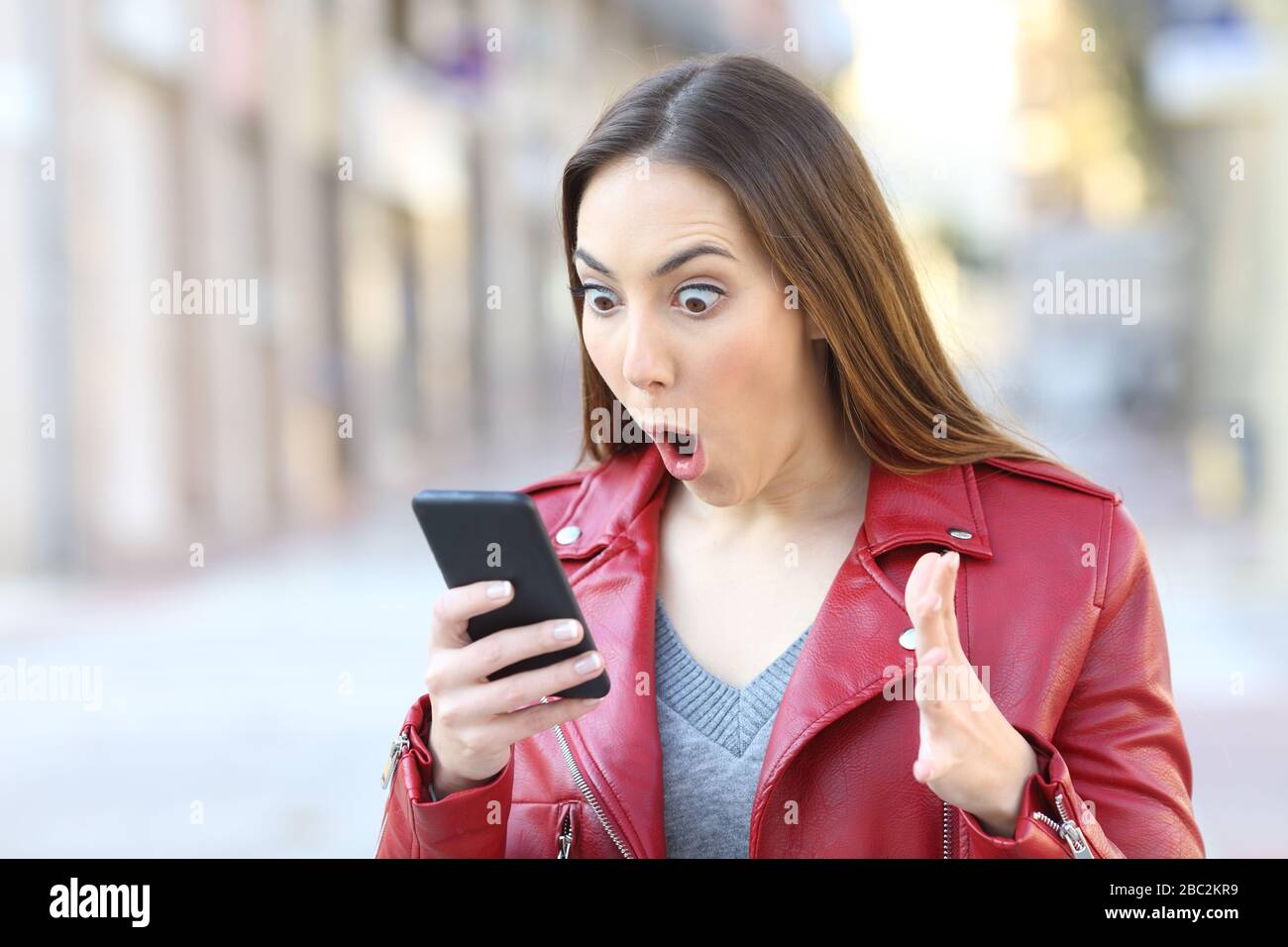 Surprised woman with eyes and mouth open wearing a red jacket looking at her phone screen Stock ...