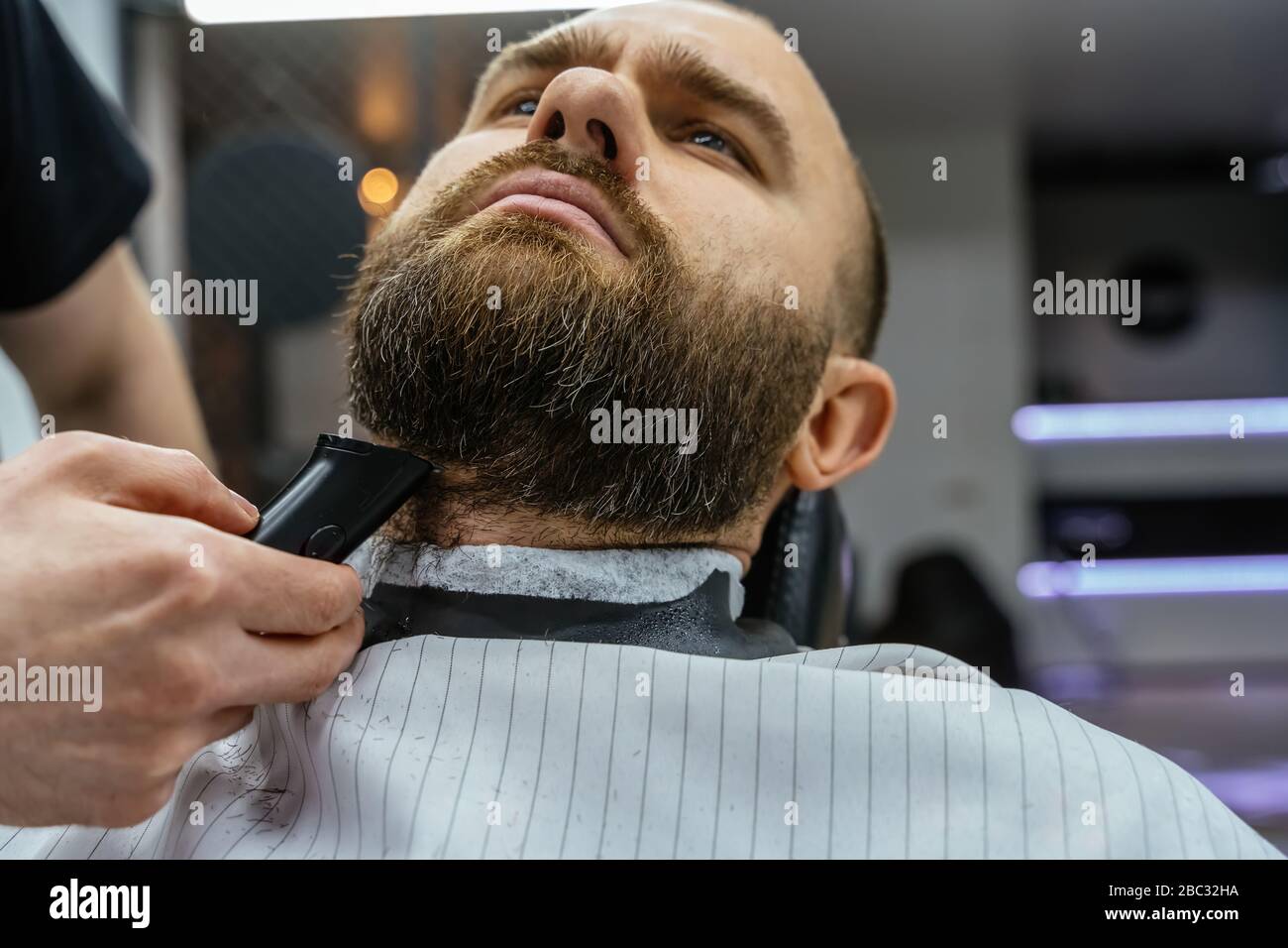 Barber trimming bearded man with shaving machine in barbershop ...