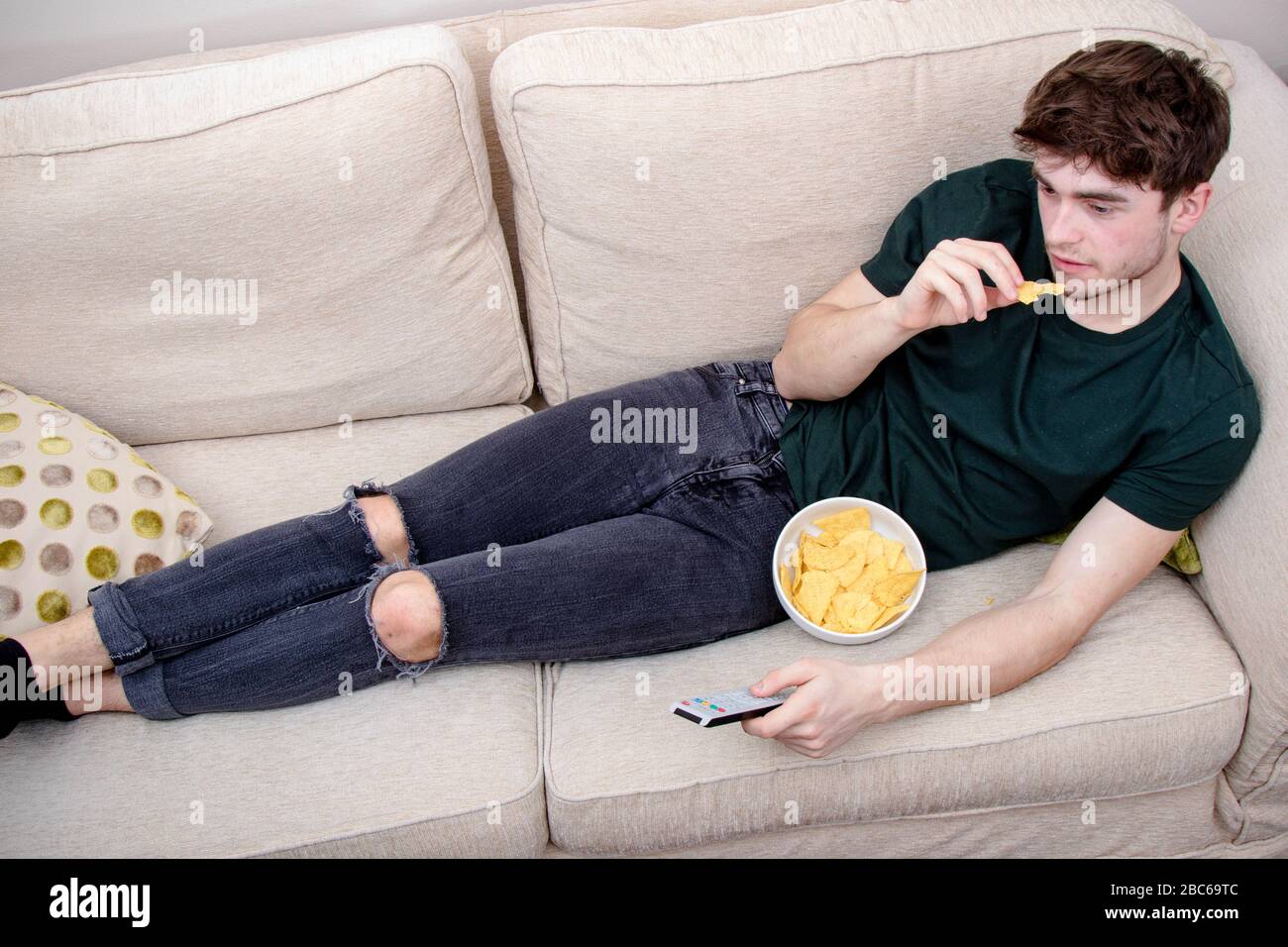 A young adult man laying on a sofa watching TV and eating snacks Stock ...