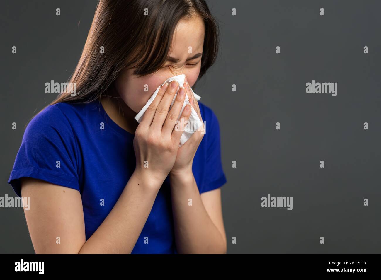 Young asian girl sneezes into tissue. Isolated woman on grey studio background is sick, has a ...