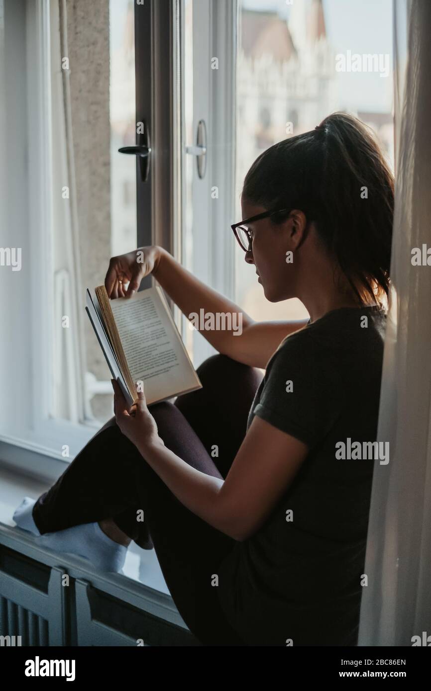 Brunette girl with glasses reading book on the window Stock Photo - Alamy