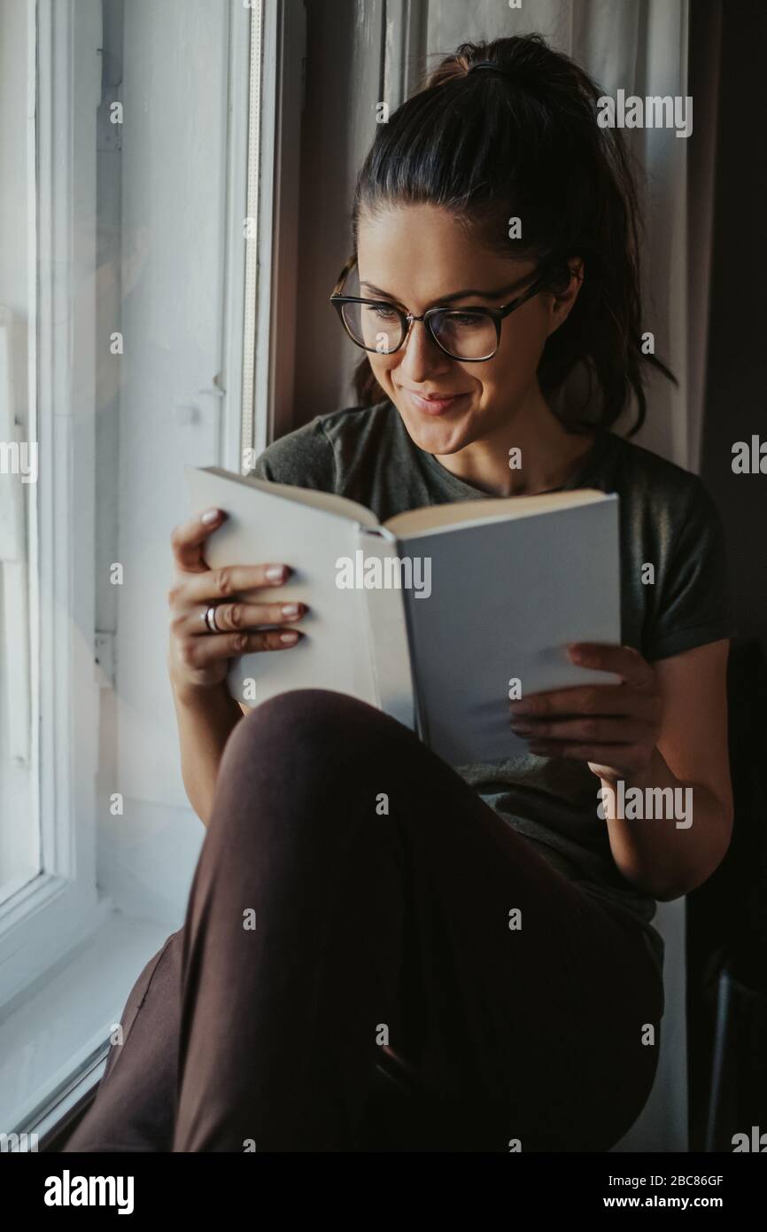 Smiling girl with glasses reading book on the window Stock Photo - Alamy