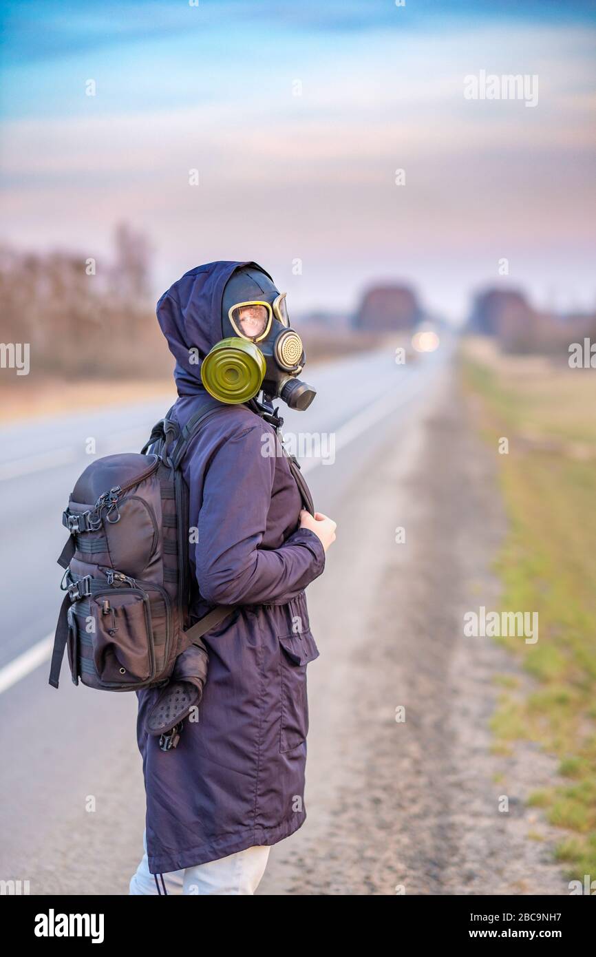 A girl in a black gas mask stands on the edge of a suburban highway ...