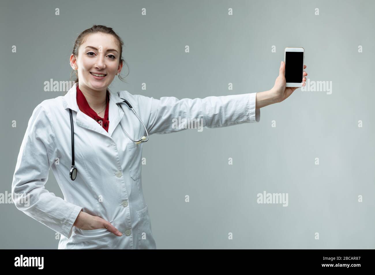 Happy friendly woman doctor holding up her smartphone in her hand with ...