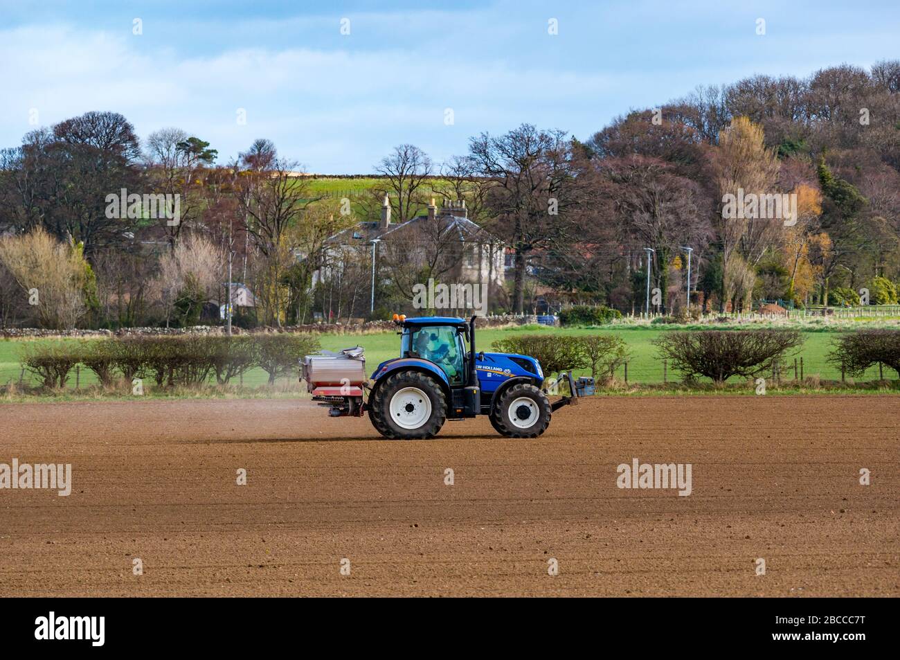 Farm tractor spreading fertiliser on a seeded field crop on a sunny ...