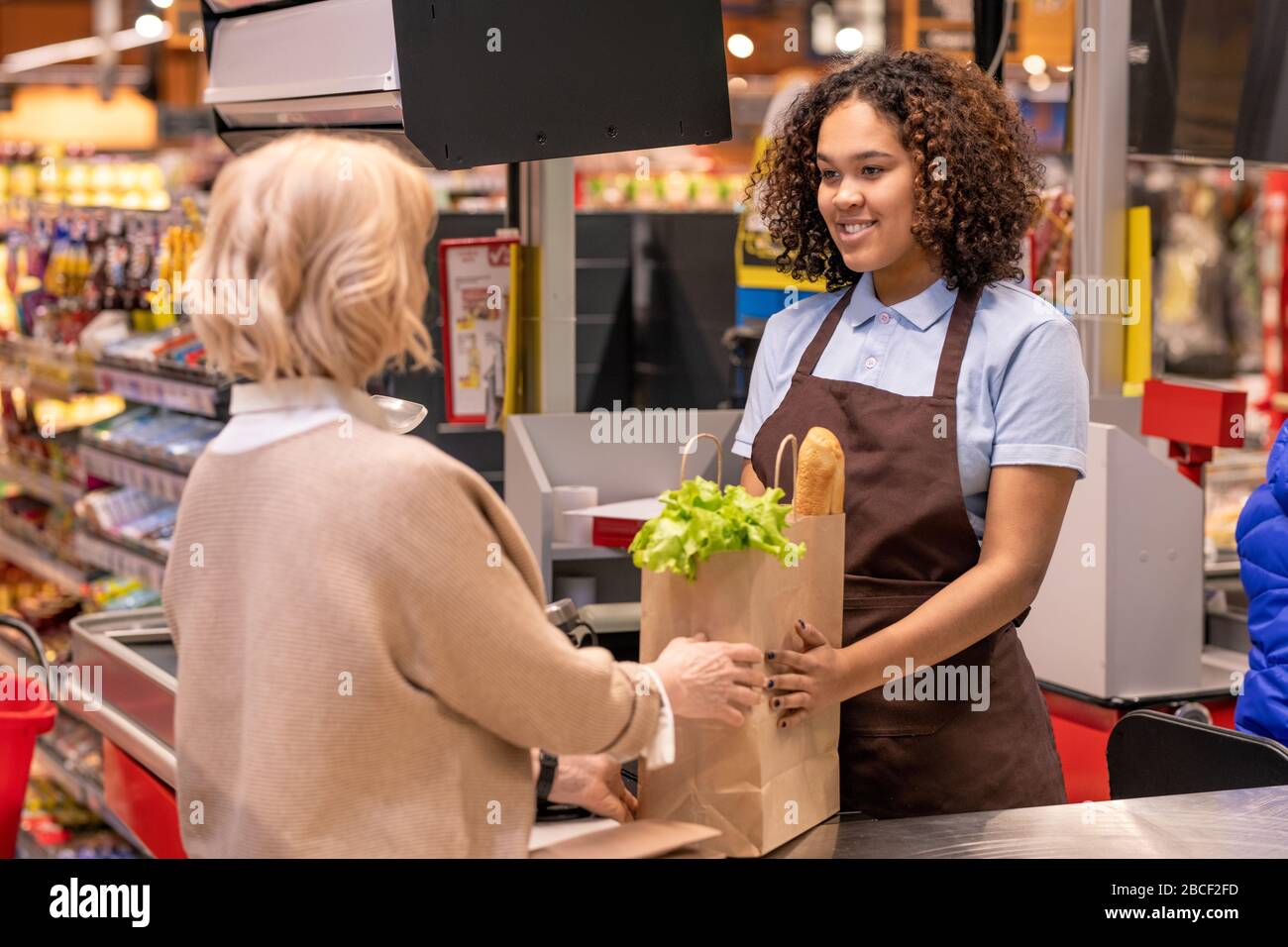 Pretty young cashier giving mature female paperbag with bread and fresh ...