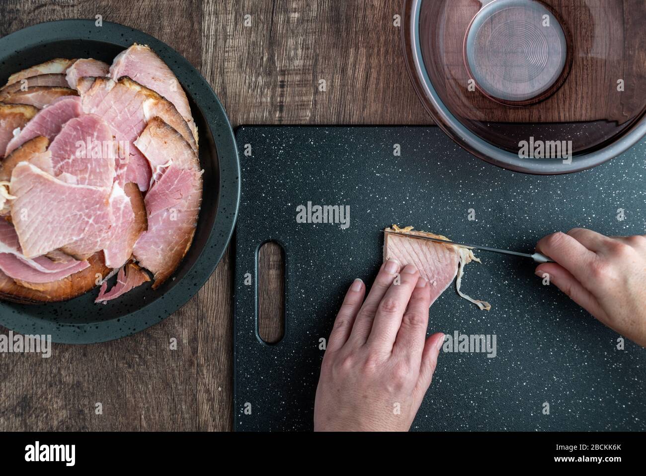 Woman’s hands using knife to slice ham, black plate stacked with ...