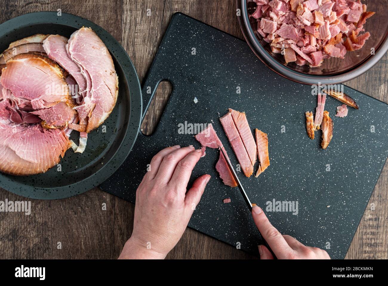 Woman’s hands using knife to slice ham, black plate stacked with ...