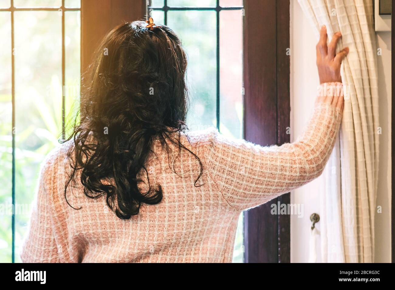 Back view of woman wear pink cardigan opens the window curtains and ...