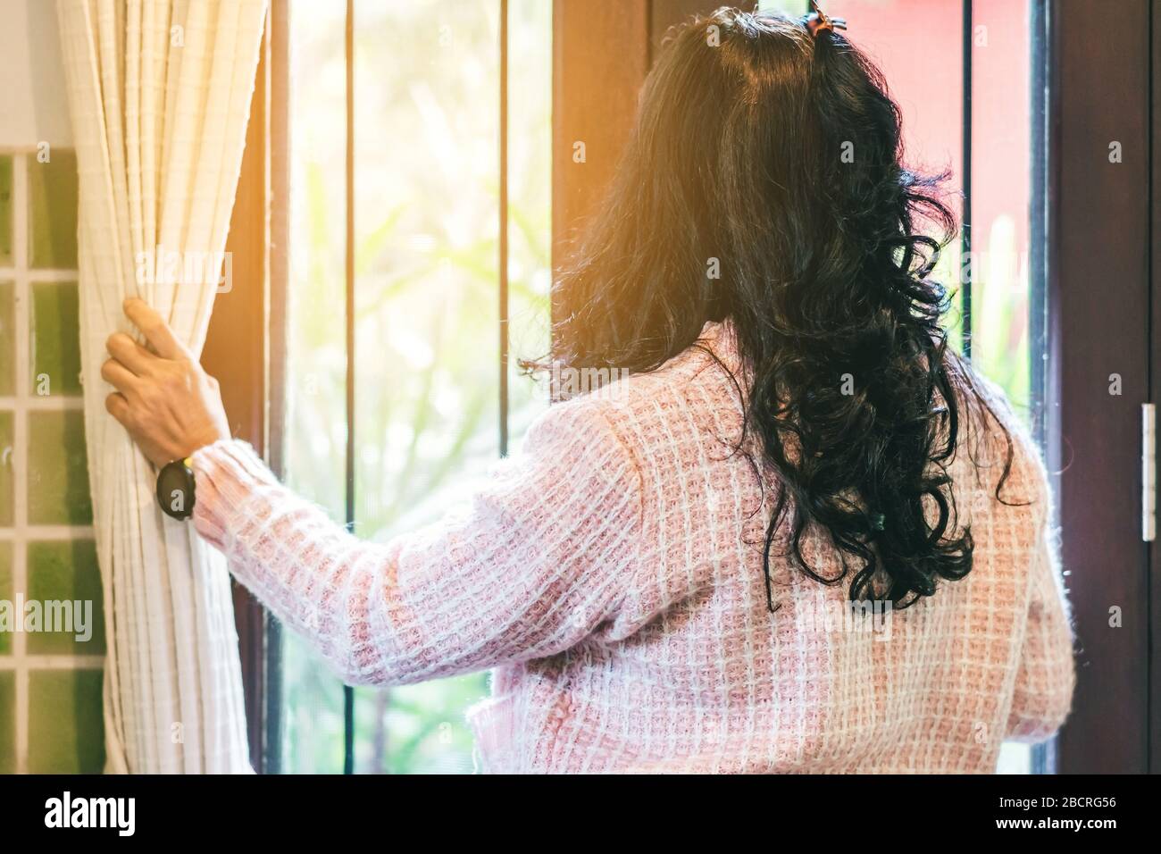 Back view of woman wear pink cardigan opens the window curtains and ...