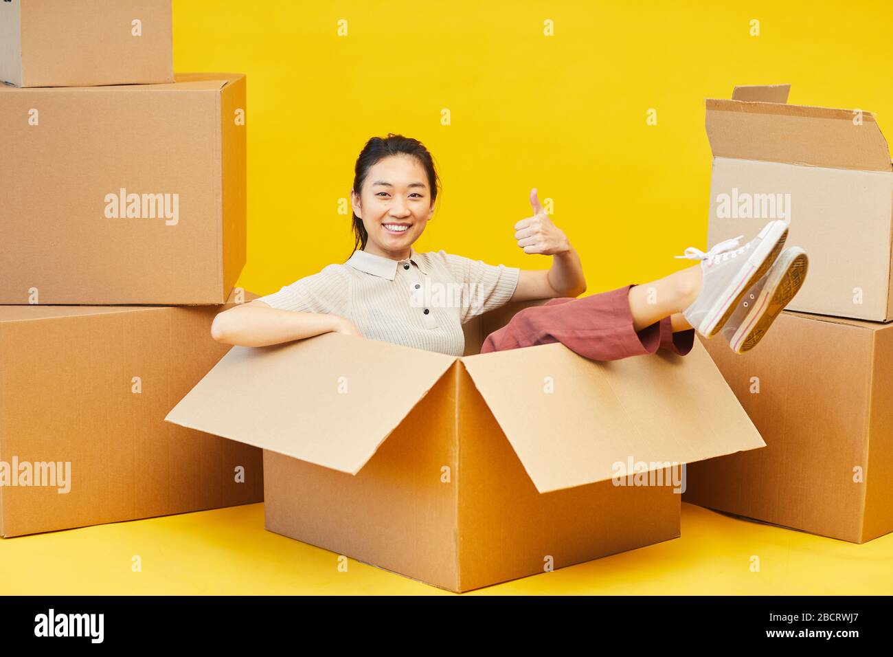 Full length portrait of happy Asian woman sitting in box and showing ...