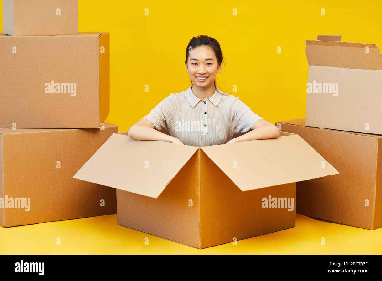 Full length portrait of happy Asian woman sitting in box and looking at ...