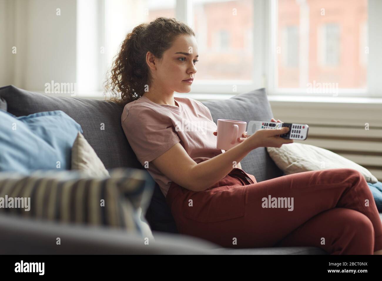 Side view portrait of young woman watching TV and holding remote while ...