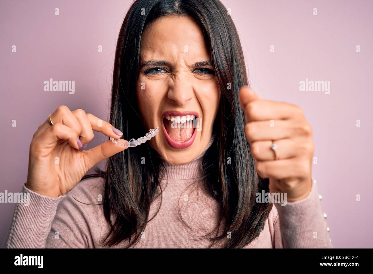 Young woman with blue eyes holding clear aligner standing over pink ...