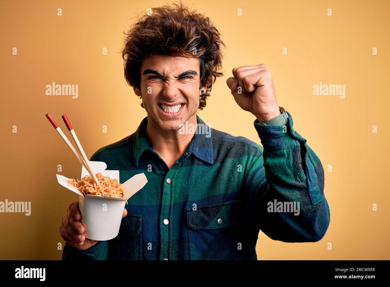 Young handsome man eating noodles standing over isolated yellow ...