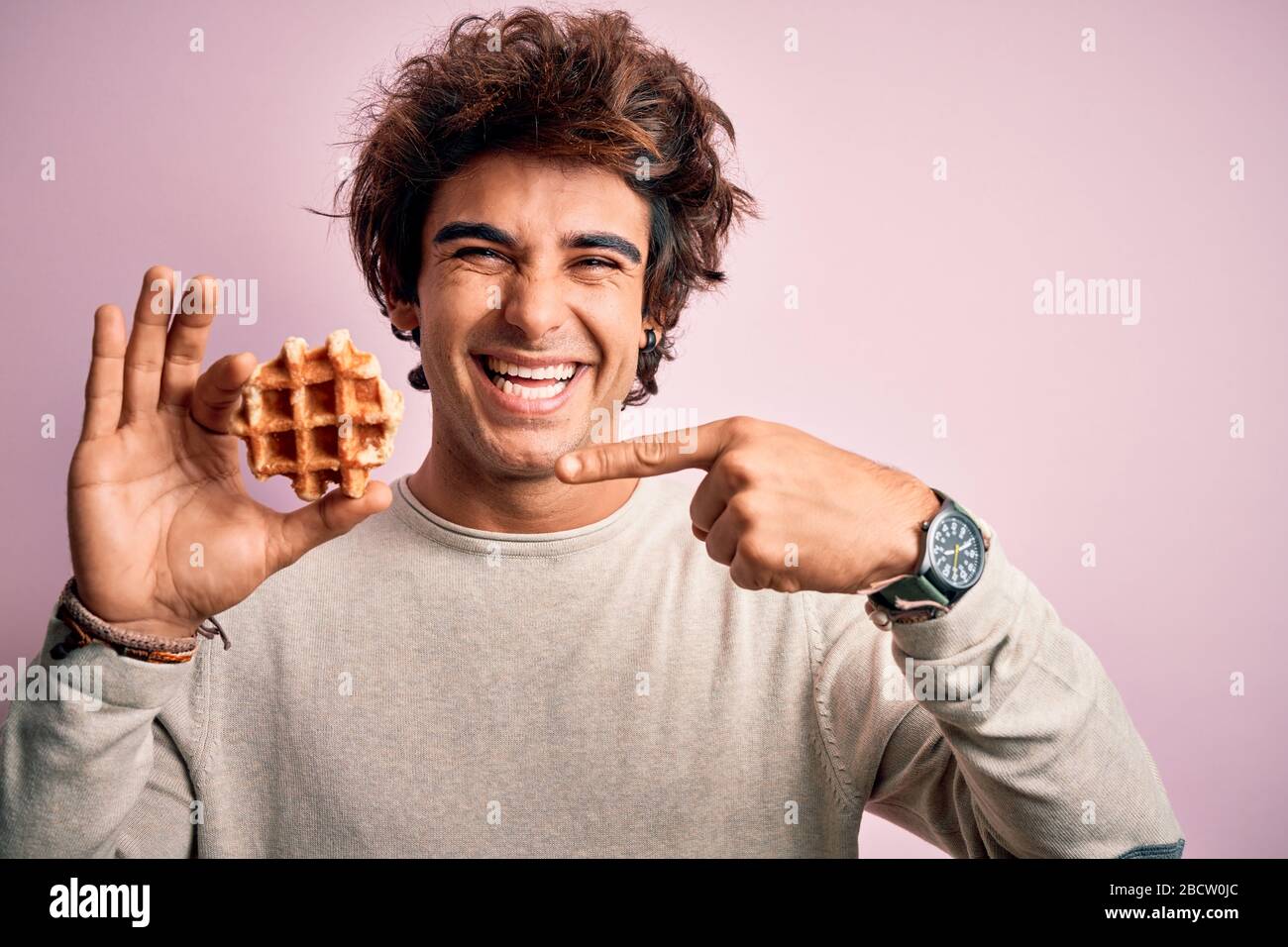 Young handsome man holding sweet waffle standing over isolated pink ...