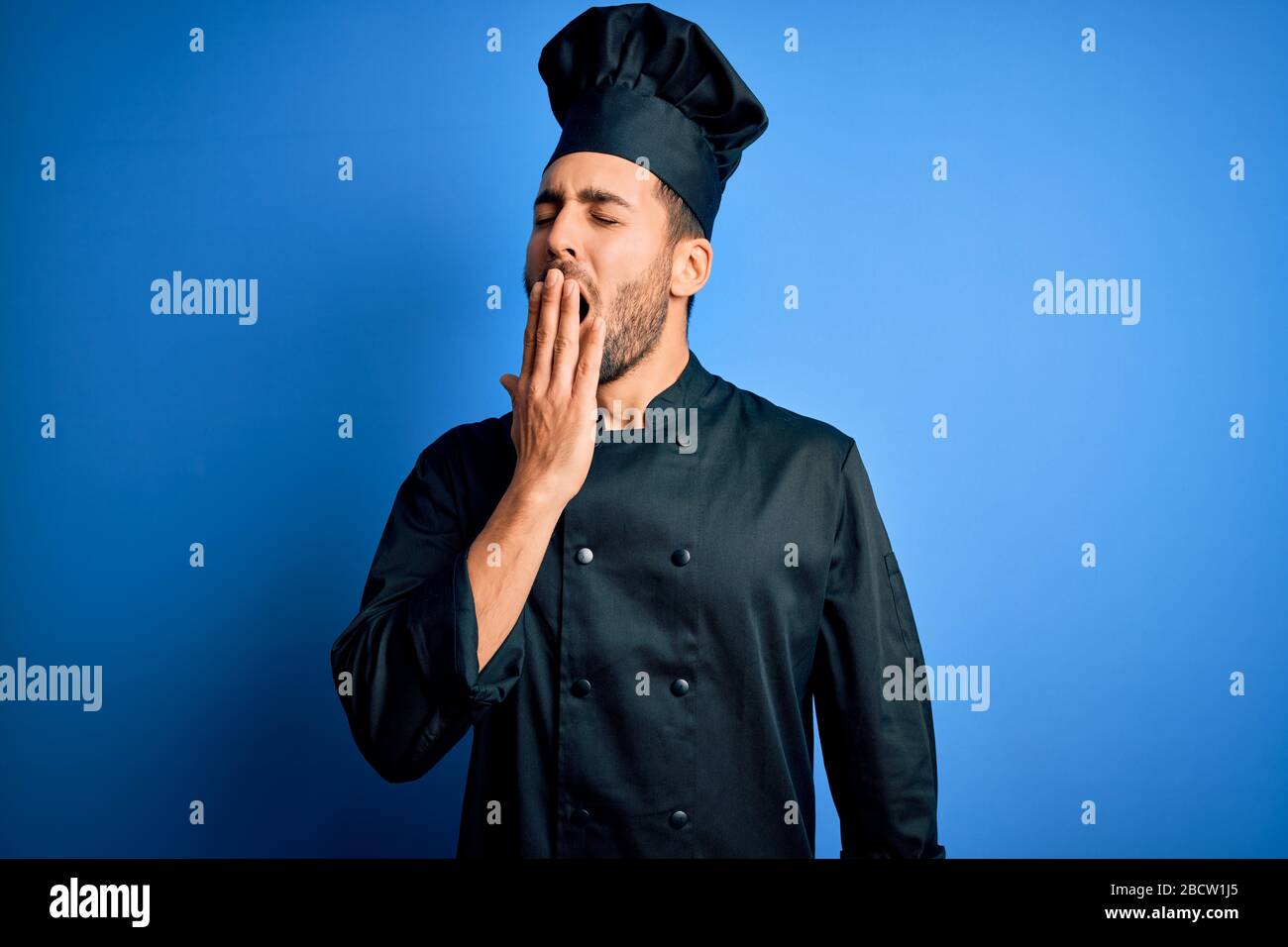 Young handsome chef man with beard wearing cooker uniform and hat over ...