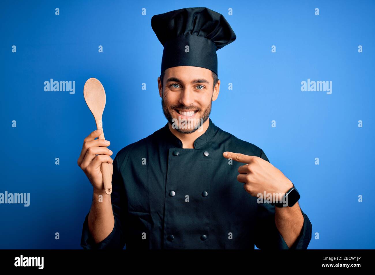 Young cooker man with beard wearing uniform holding wooden spoon over ...