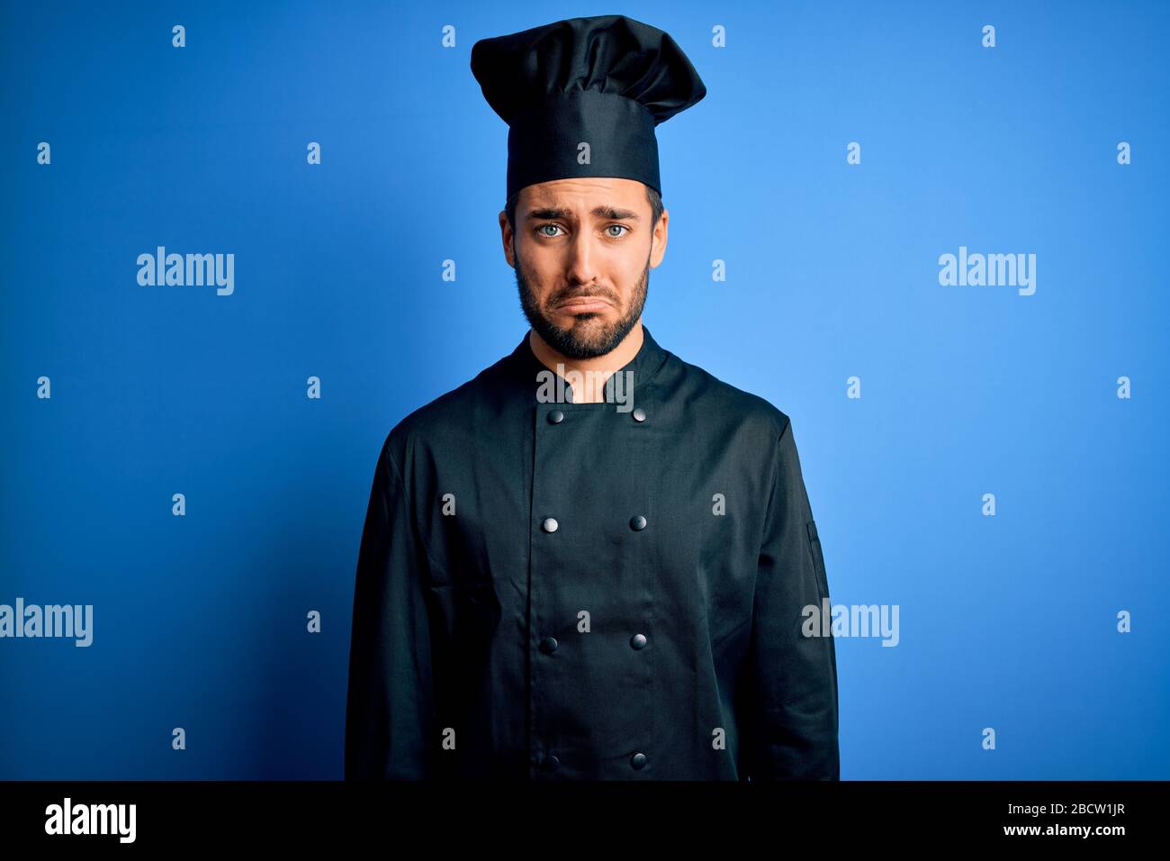 Young handsome chef man with beard wearing cooker uniform and hat over ...