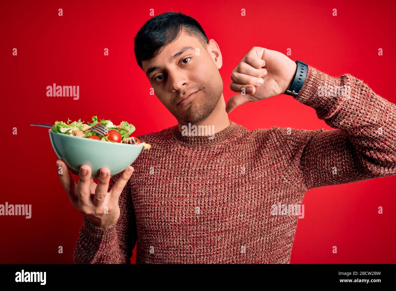 Young hispanic man eating vegetarian healthy salad over red isolated ...