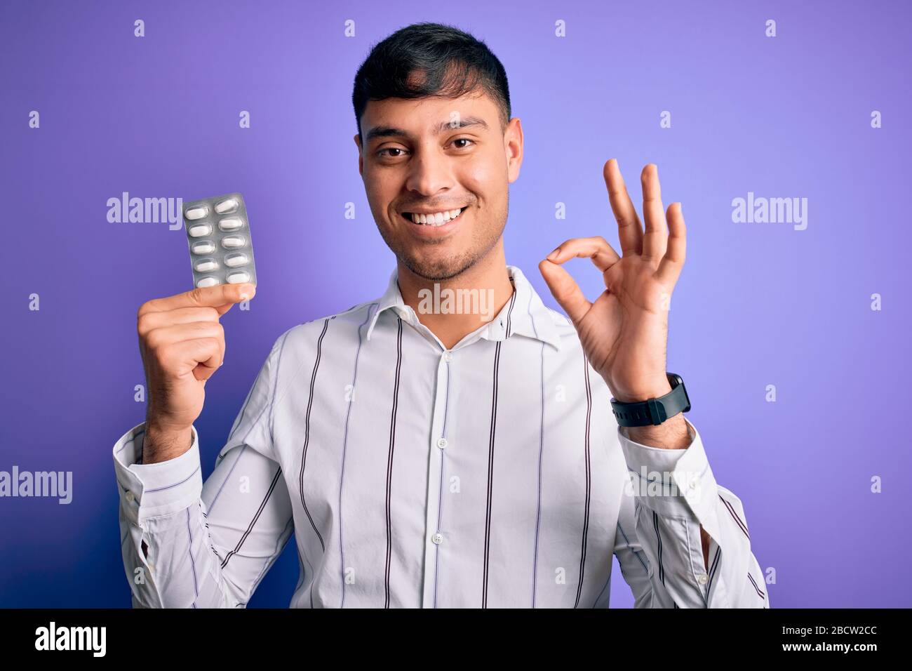 Young hispanic man holding pharmaceutical antibiotics pills over purple ...