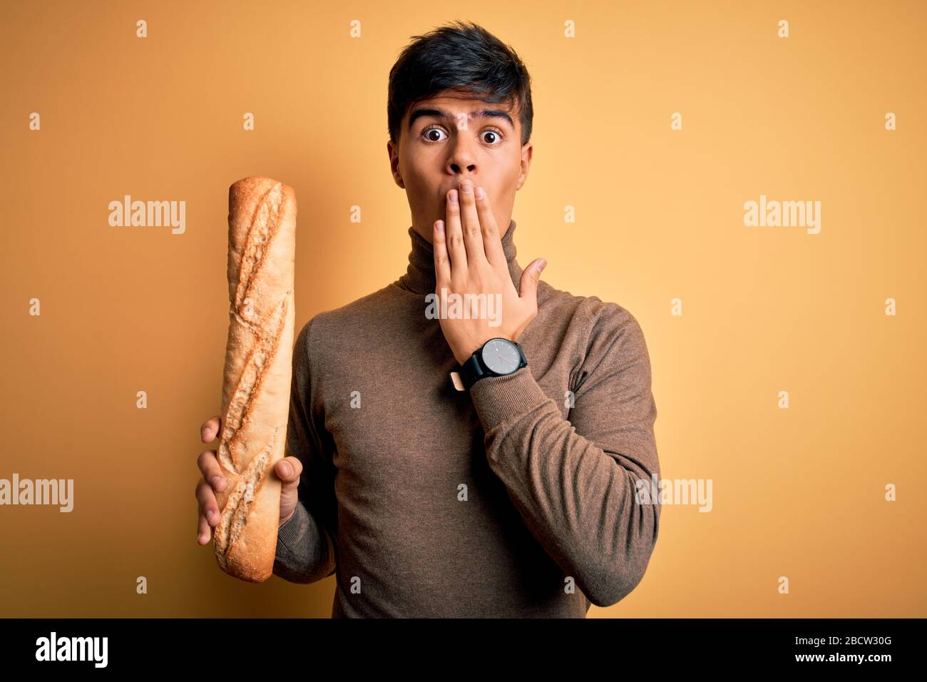 Young handsome man holding homemade fresh bread over isolated yellow ...