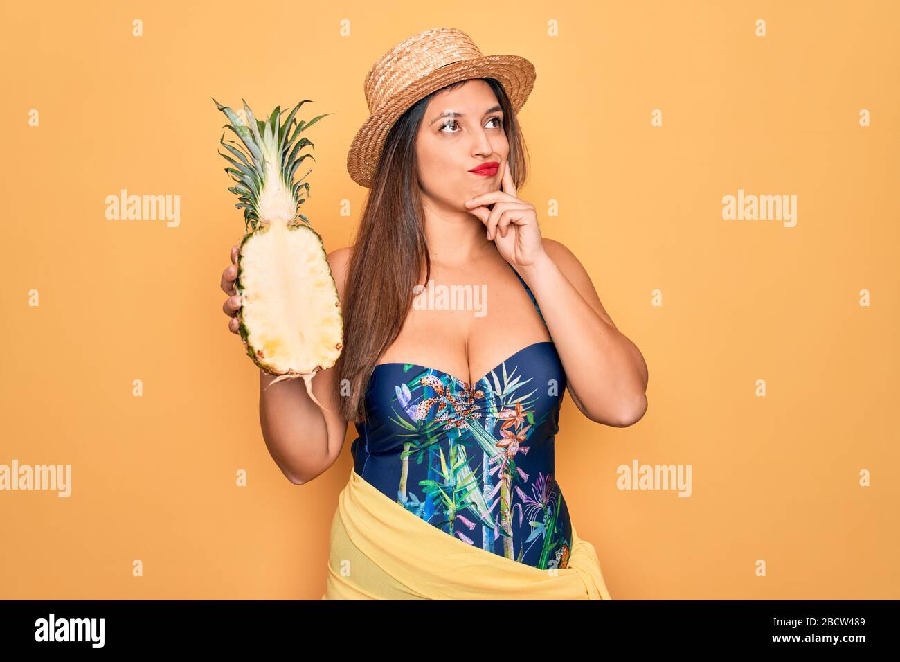 Young hispanic woman wearing summer hat and swimsuit holding tropical pineapple serious face ...