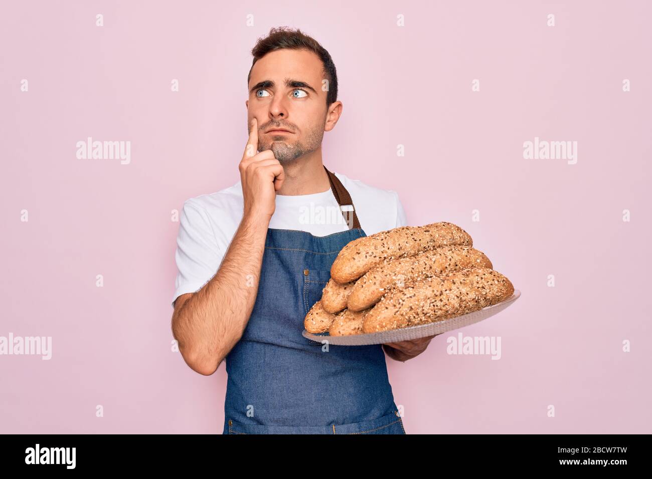 Young handsome baker man with blue eyes wearing apron holding tray with ...