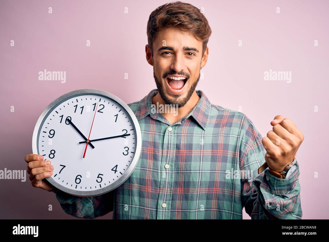 Young man with beard doing countdown using big clock over isolated pink ...