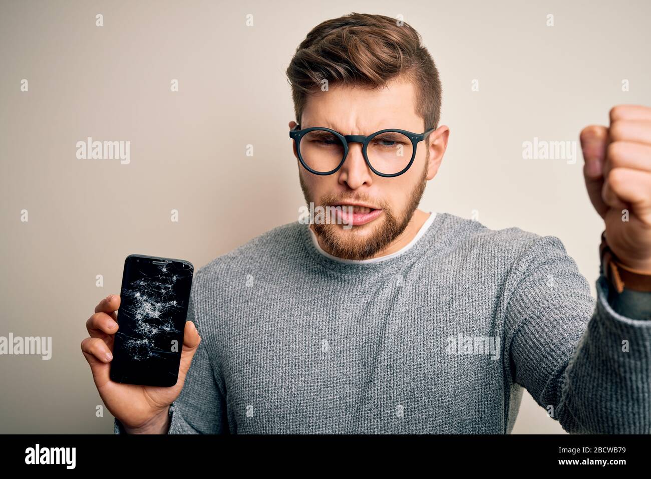 Young blond man with beard and blue eyes wearing glasses holding broken ...