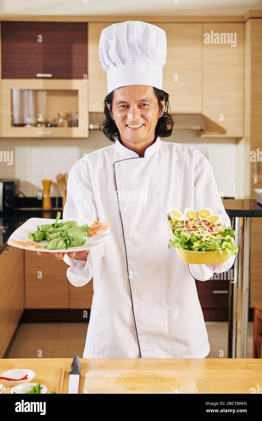 Smiling happy Asian chef standing in kitchen and holding two plates ...