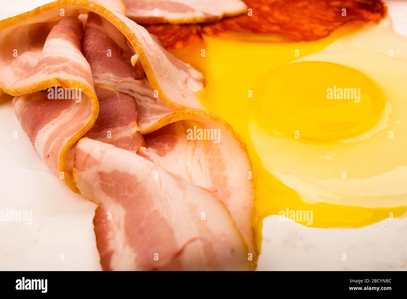 a broken chicken egg without a shell and slices of bacon on a white ...